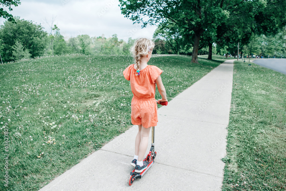 Young girl child in red orange romper riding scooter on street road ...