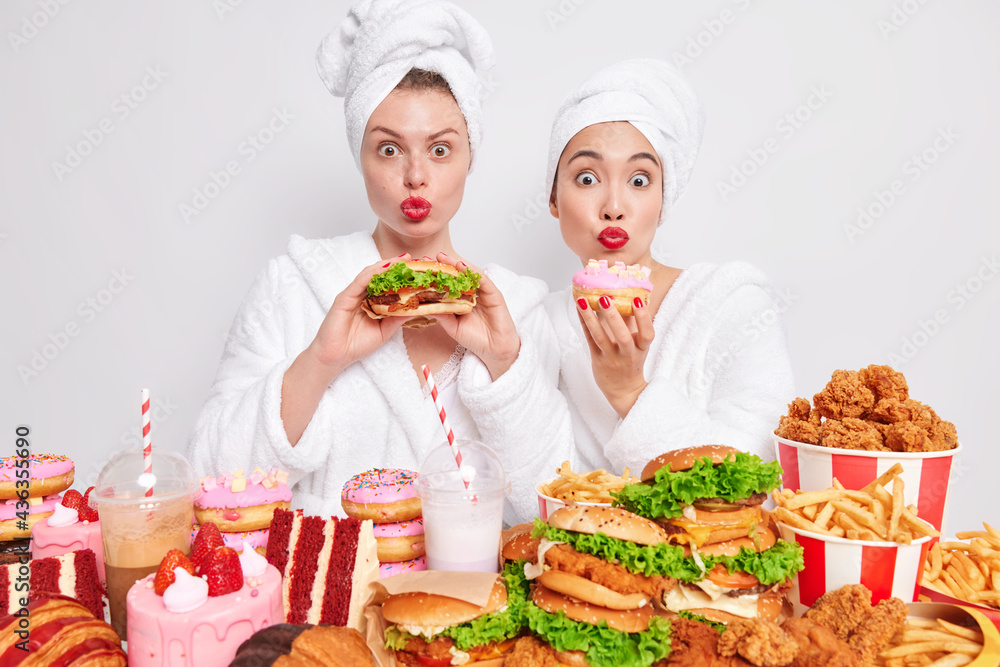 Never enough. Two hungry female models in robes consume junk food keeps ...