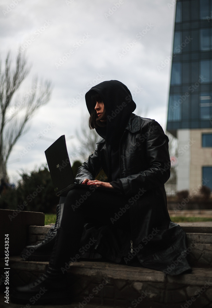Girl in black leather coat and hood with laptop on city street, concept ...