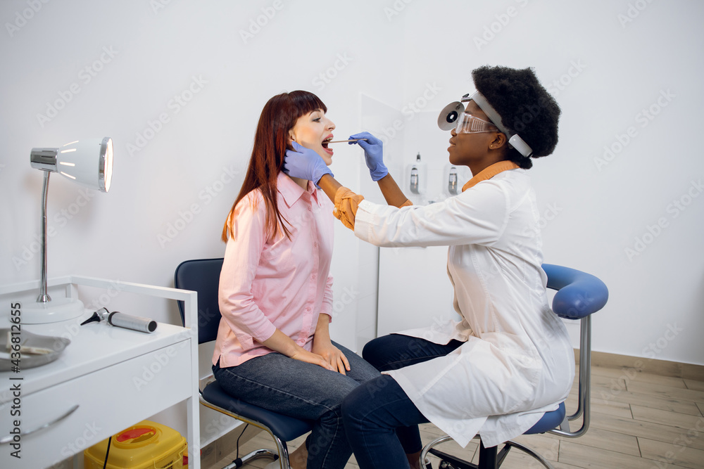 Young high skilled female afro ENT doctor checking woman's throat ...