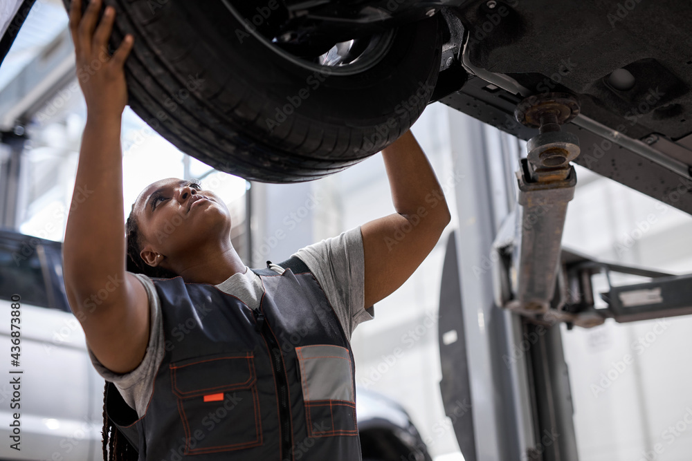 African female auto mechanic changing wheel tire in car in garage, side view. Beautiful black ...