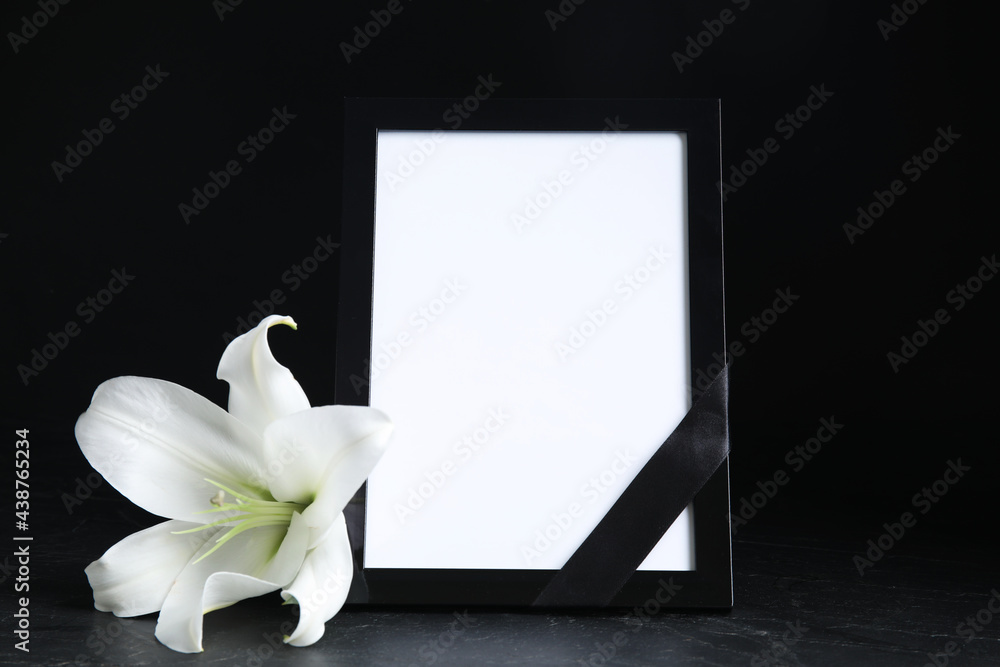 Funeral photo frame with ribbon and white lily on black table against ...