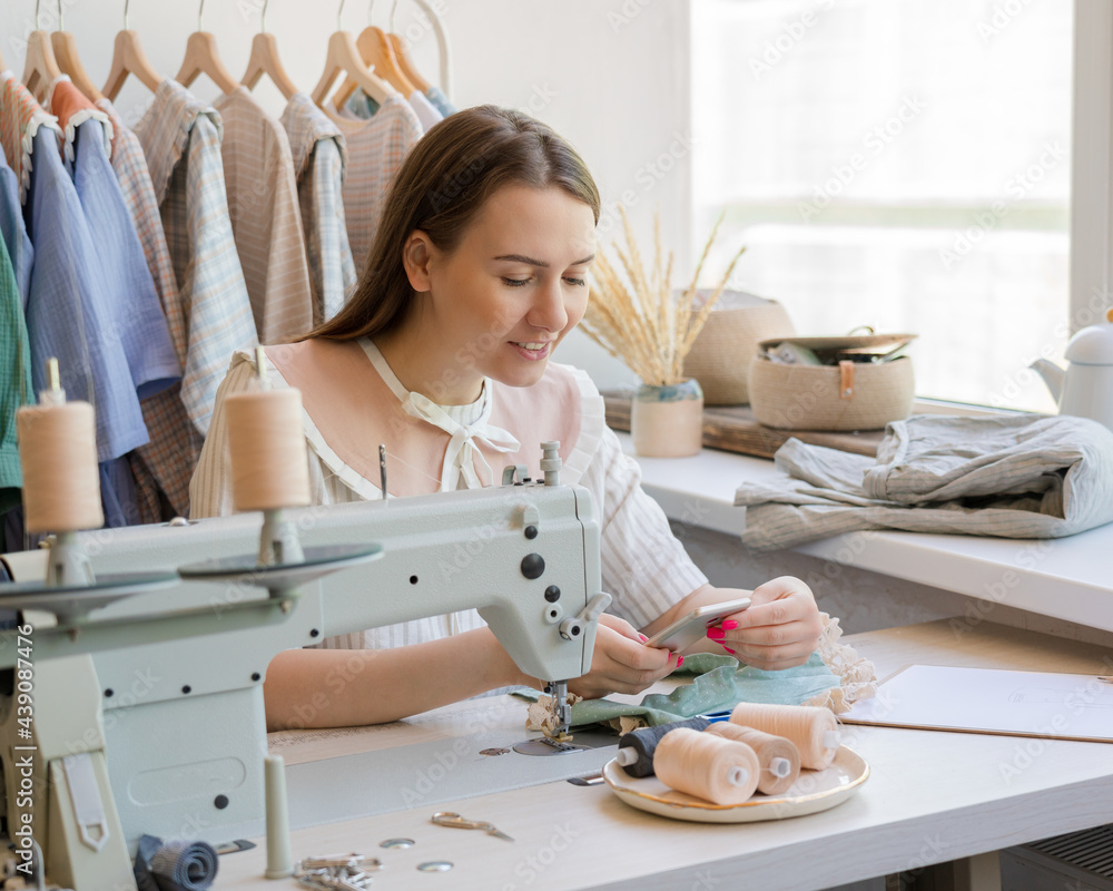 Happy young woman dressmaker or seamstress with cell phone at workplace ...