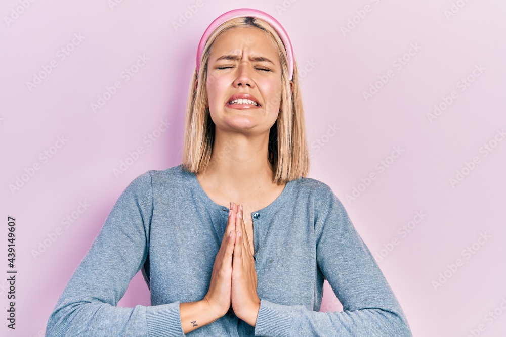 Beautiful blonde woman standing over pink background begging and ...