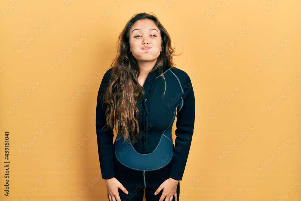 Young hispanic girl wearing diver neoprene uniform puffing cheeks with ...