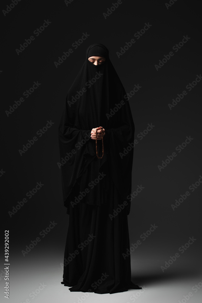 full length view of muslim nun in black niqab and abaya praying with ...