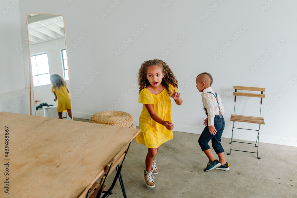 Young biracial brother and sister run around room with wood table and ...
