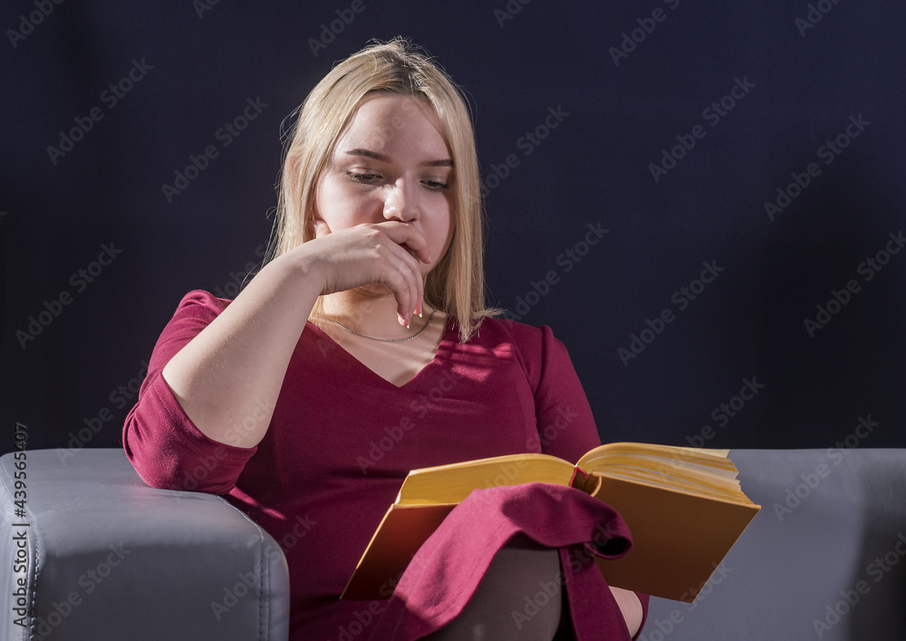 Sad blonde girl sitting on couch and reading book. Woman is resting and ...