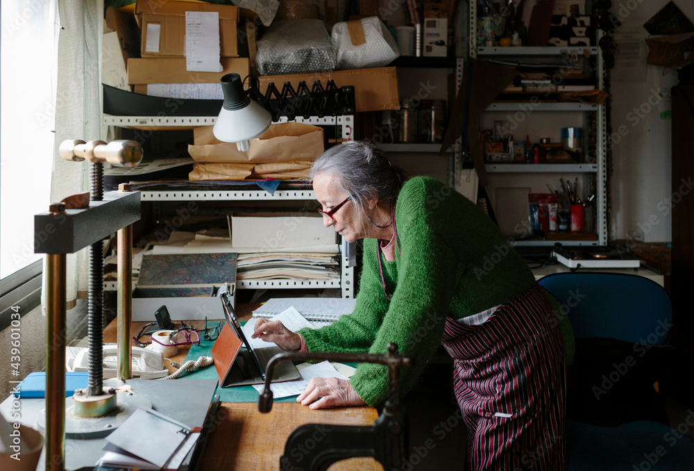 Grey-haired artisan woman using ipad in her small bookbinding workshop ...