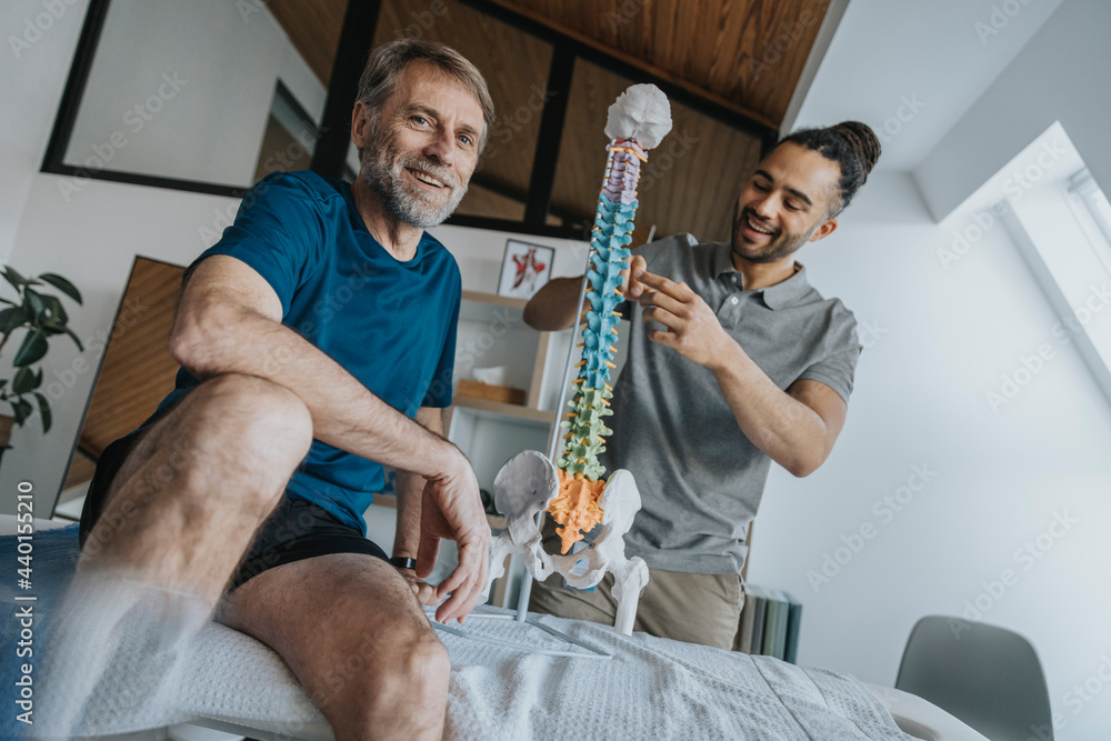 Smiling male physical therapist explaining spine model by patient in ...
