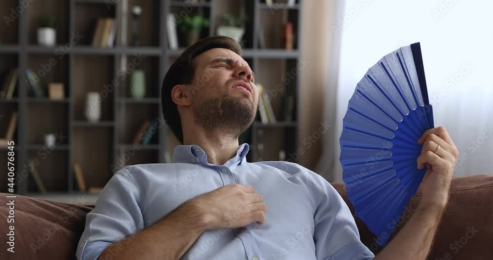 Tired overheated young man cooling himself wave hand and handheld fan ...
