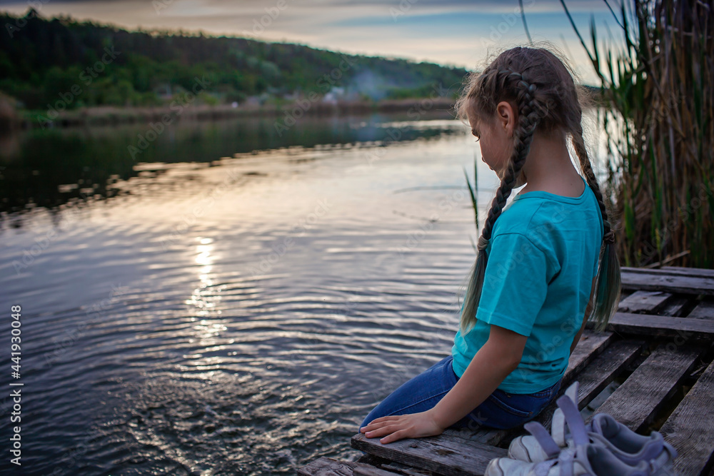 Cute preteen girl sits on a wooden pier by the lake and stirs feet in ...