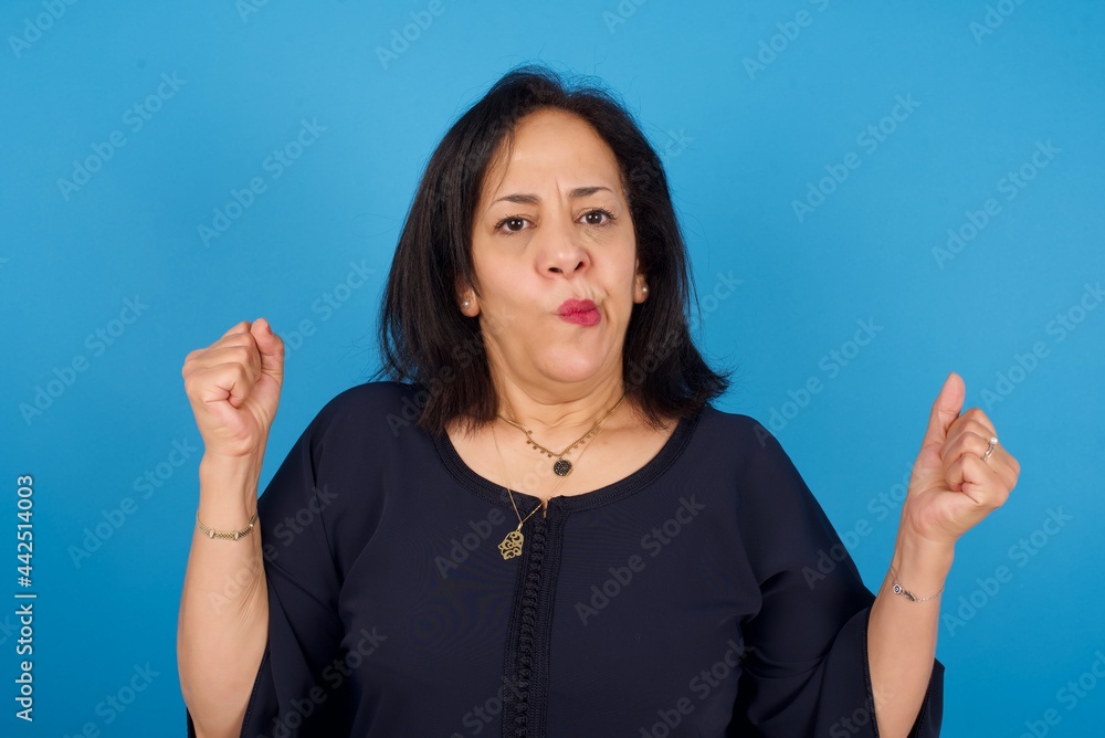 Irritated middle aged Arab woman standing against blue background blows ...