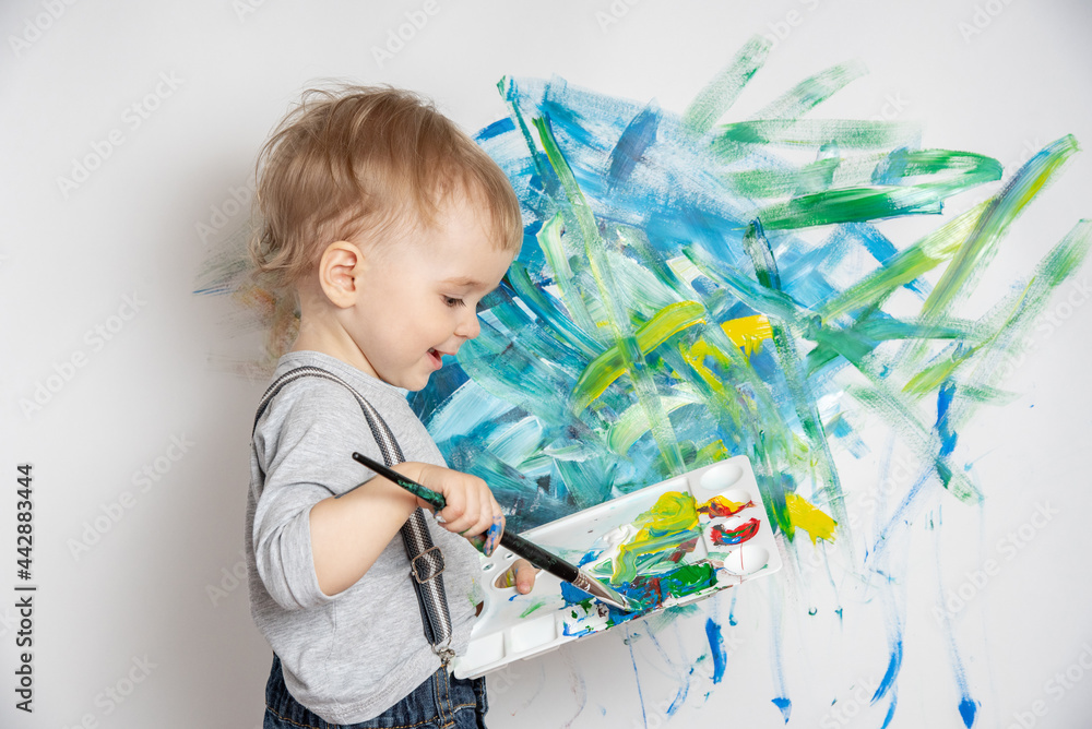 Little child with a brush and a palette of paints in his hands draws on ...