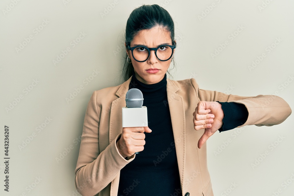 Young hispanic woman holding reporter microphone with angry face ...