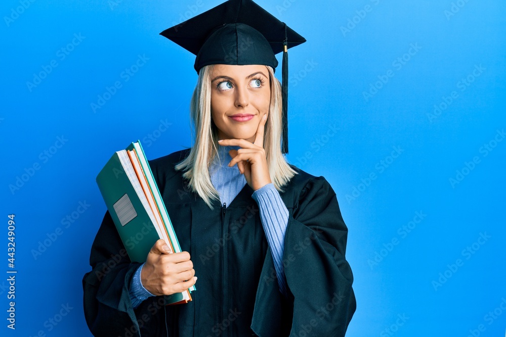 Beautiful blonde woman wearing graduation cap and ceremony robe holding ...