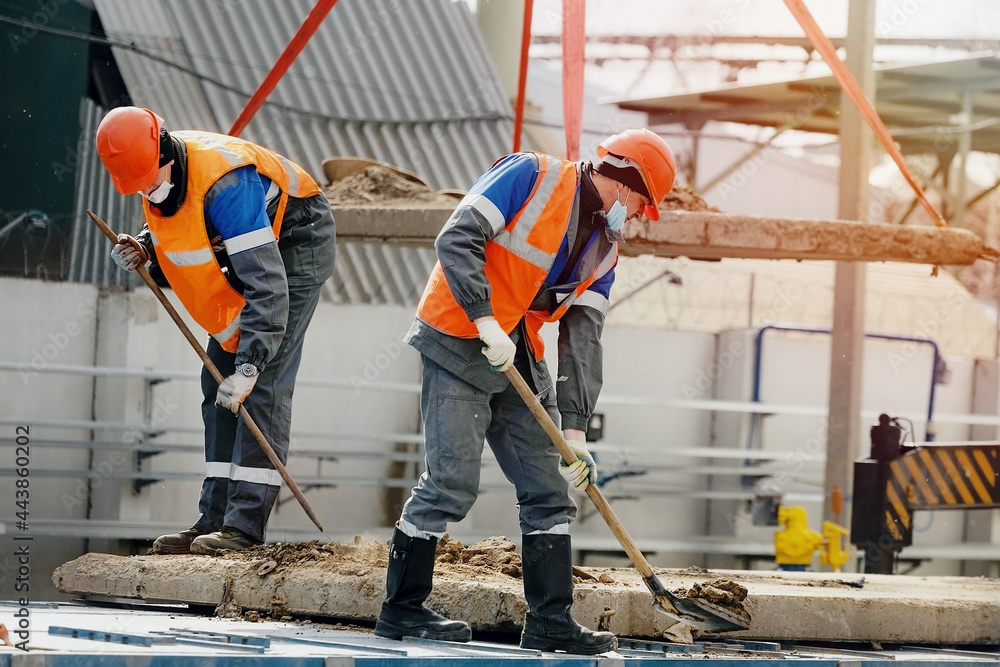 Two workers in hard hats, work clothes and a medical mask work with ...