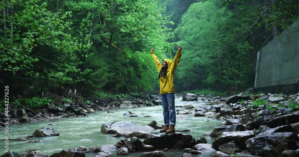 Emotional young woman traveler in yellow raincoat standing on the rock ...