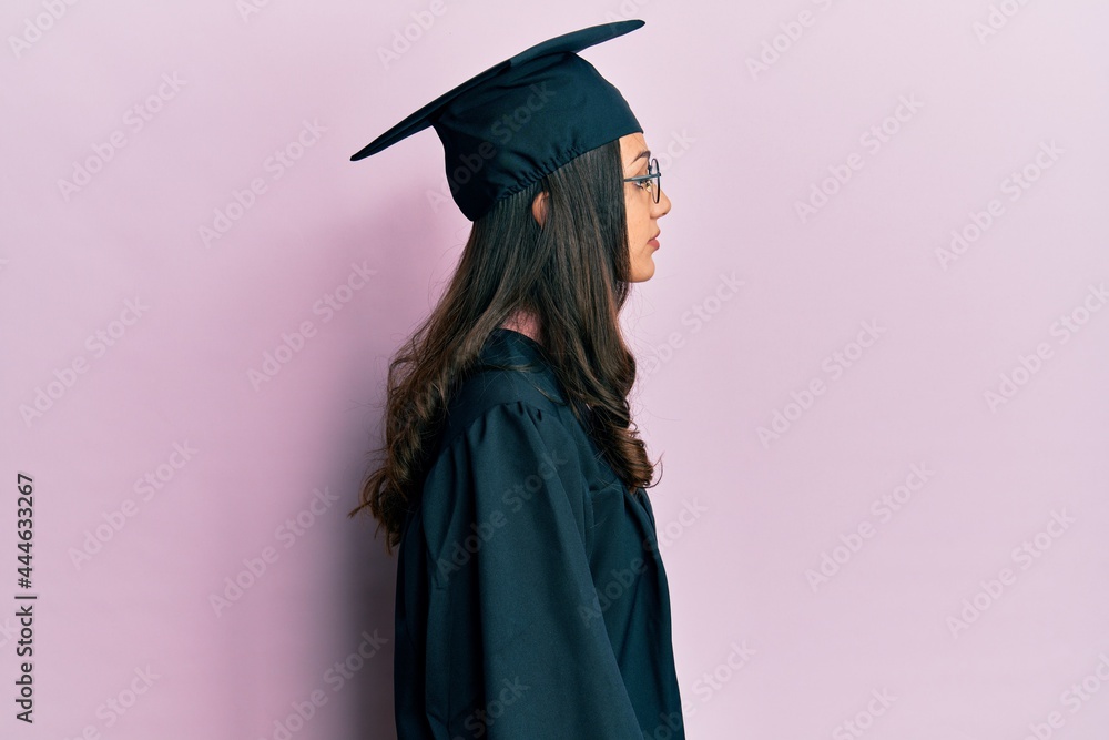 Young hispanic woman wearing graduation cap and ceremony robe looking ...