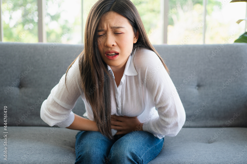 Unhappy Asian woman sitting on the sofa and holding on stomach ...