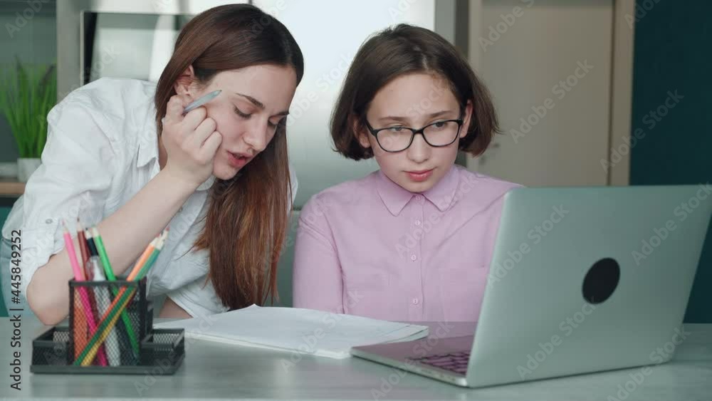 Serious daughter doing homework next to mother in front computer at ...