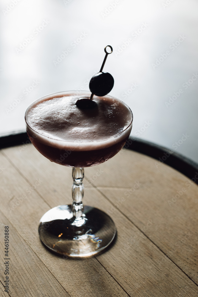 frothy cocktail in vintage coupe glass on barrel with black cherry on ...