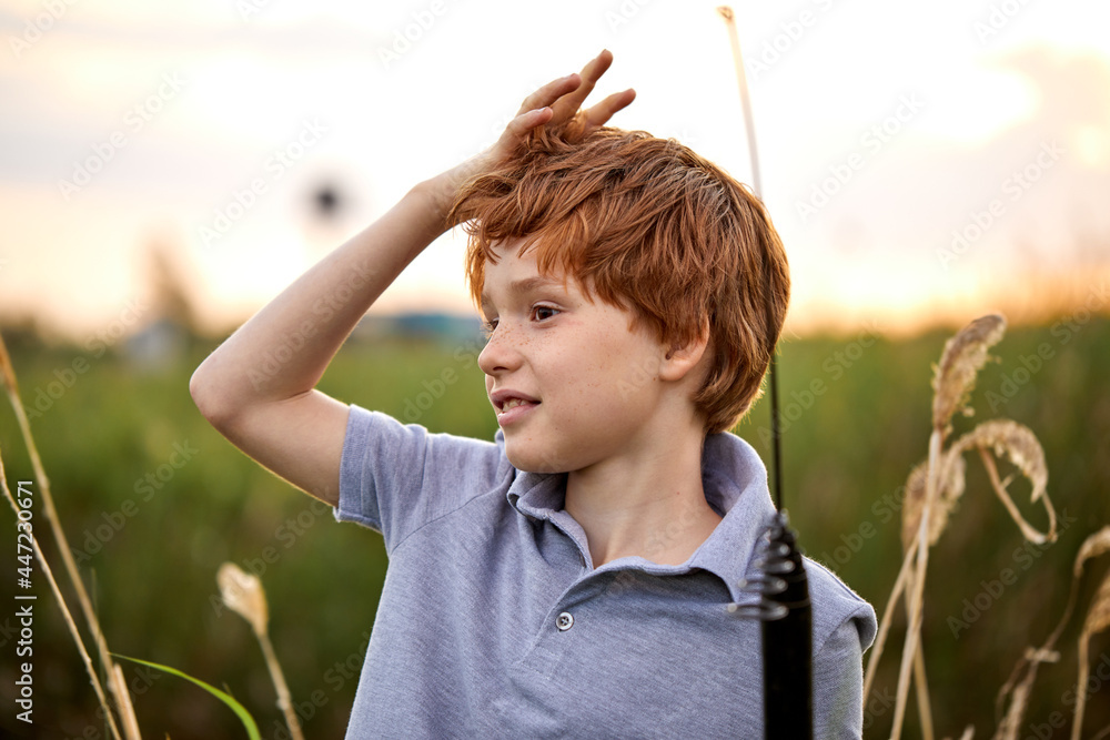 Cute smiling redhead kid boy looking at side, standing in summer field ...