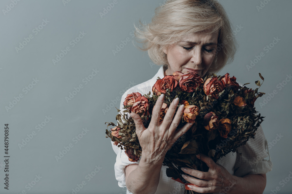 Sad elderly woman touching bouquet of withered rose flowers. Gray ...