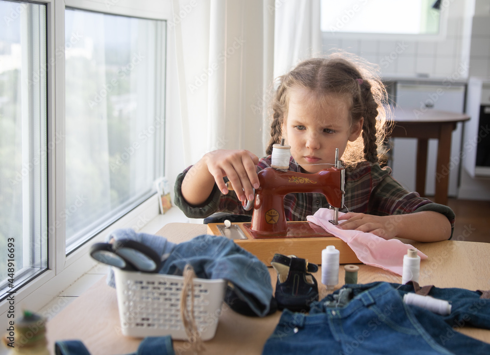 A little girl sews clothes for dolls on a toy sewing machine, a child ...