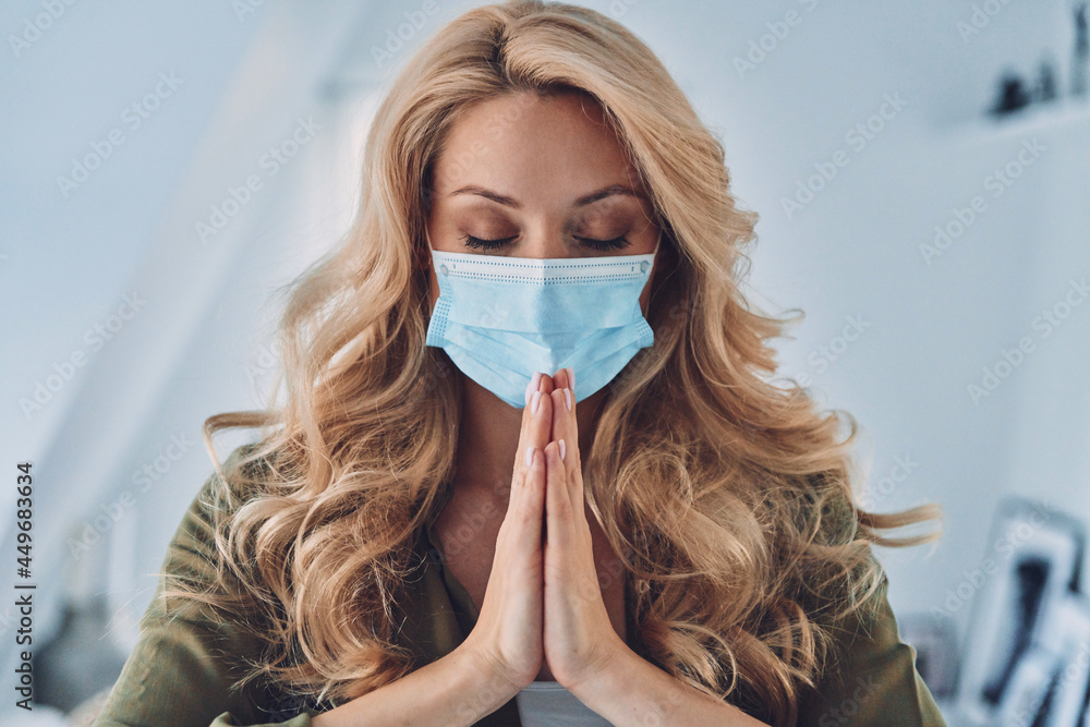 Beautiful young woman in protective face mask praying while keeping ...
