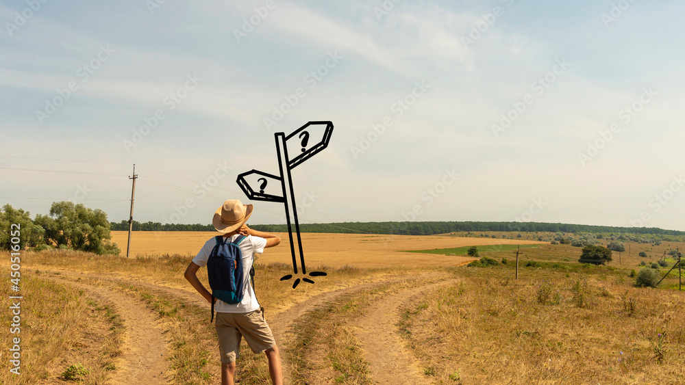 A young boy with a backpack stands at a fork in two roads at the road ...