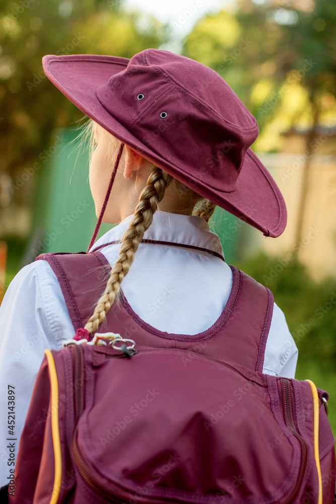 A girl wearing school uniform, white shirt, maroon backpack and a hat ...
