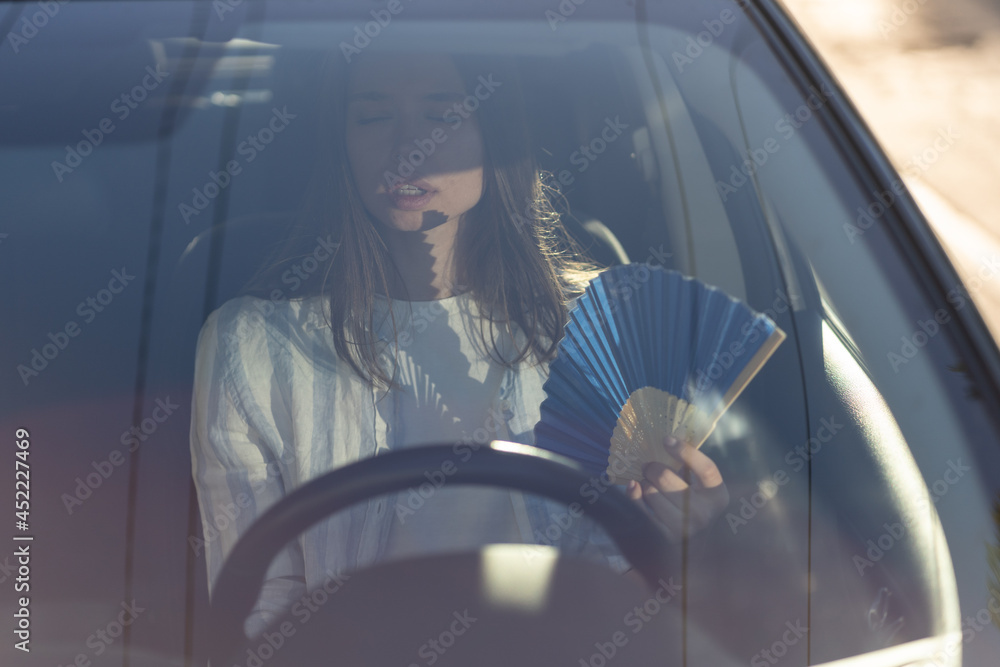 Young girl suffer from heat inside car waving fan for cooling air with ...