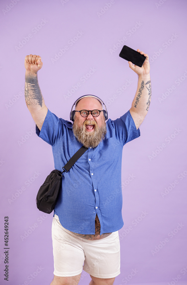 Excited middle aged fat man with headphones and crossbody bag raises up ...