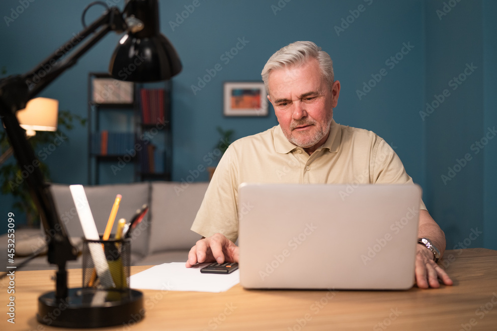 A concerned older man wonders as he sits at his laptop in his living ...
