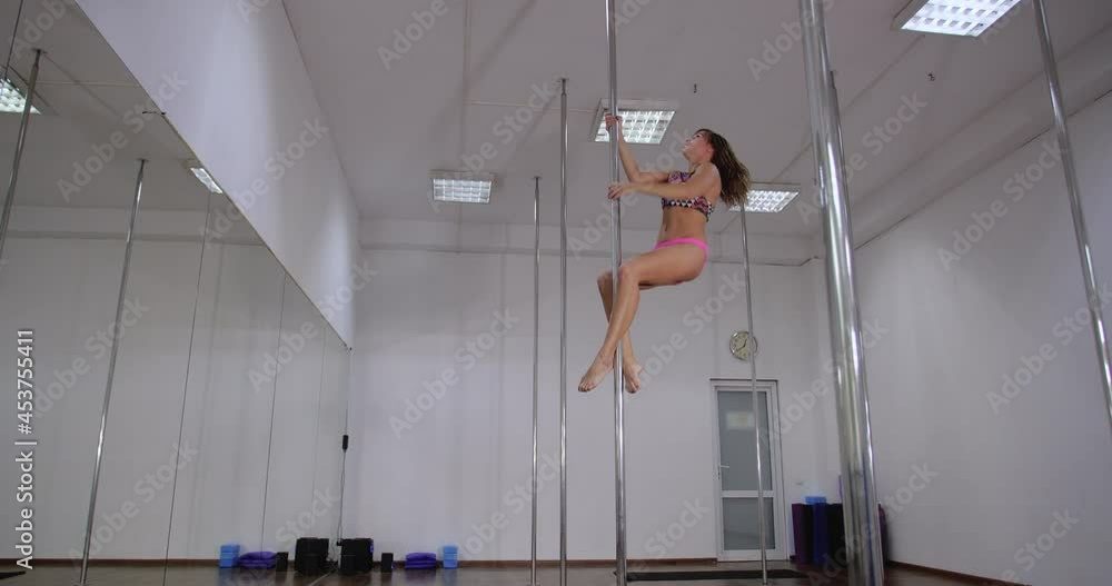 Attractive sporty young woman in pink clothes perform exercises on a pole in a pole-dance studio ...