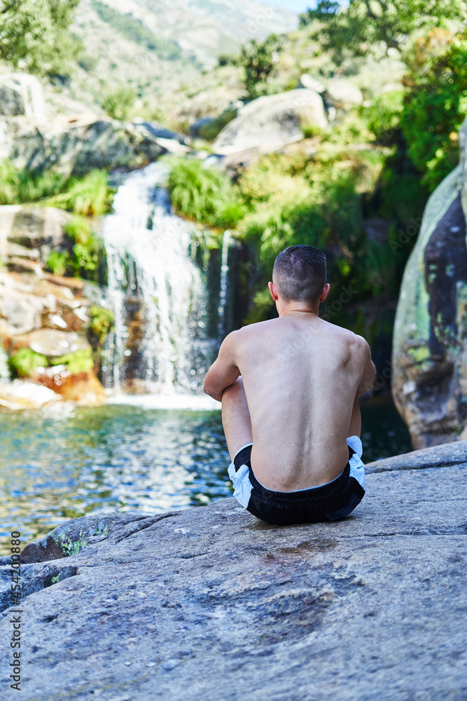 Young boy sitting on his back on a rock looking at the river with ...