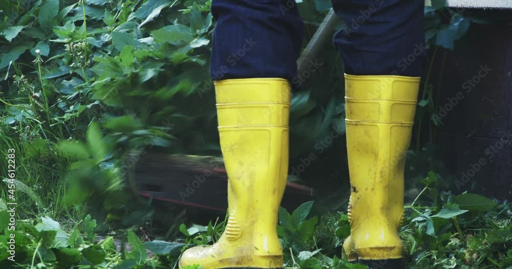 Close up of gardener in yellow rubber boots mowing high grass using ...