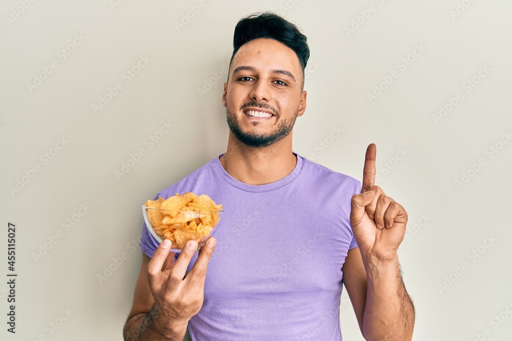 Young arab man holding potato chip smiling with an idea or question ...