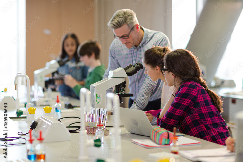 Male teacher helping girl students using microscope, conducting ...