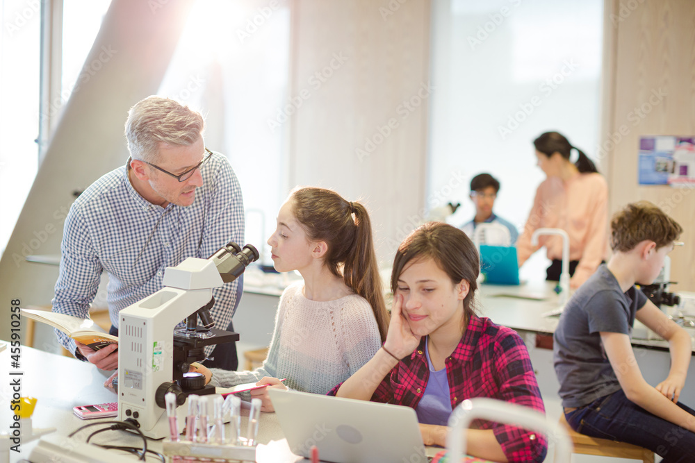 Male teacher helping girl students using microscope, conducting ...