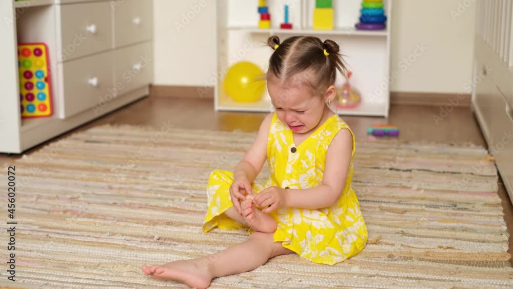 A two-year-old girl in a dress is crying, fingering her toes, sitting ...