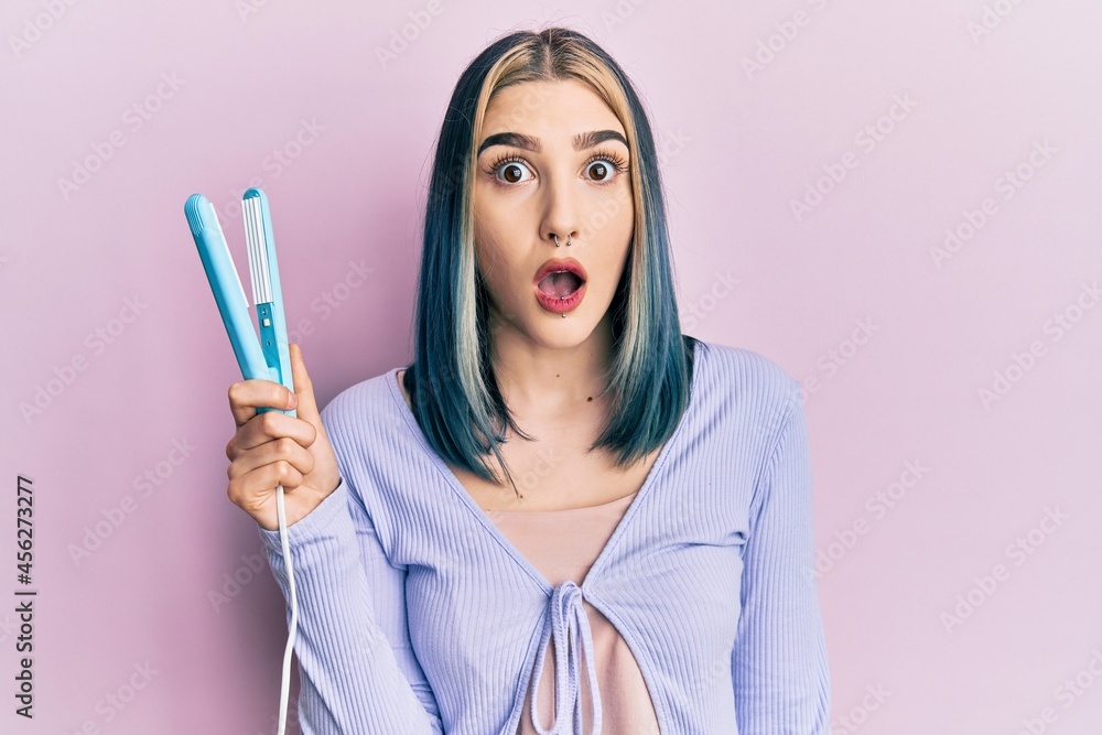 Young modern girl holding hair straightener scared and amazed with open ...