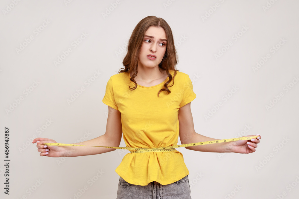 Portrait unhappy smiling woman in yellow T-shirt measuring her waist ...