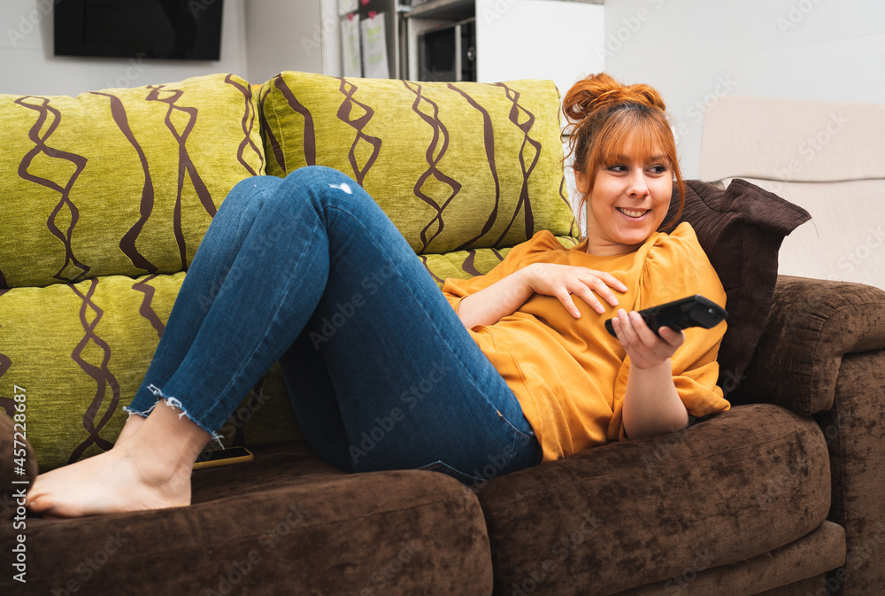 Caucasian female lying on a couch changing the channel with a remote ...