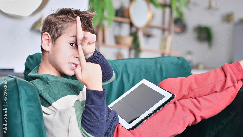 Cheerful little boy smiling while sitting on couch and using tablet at ...