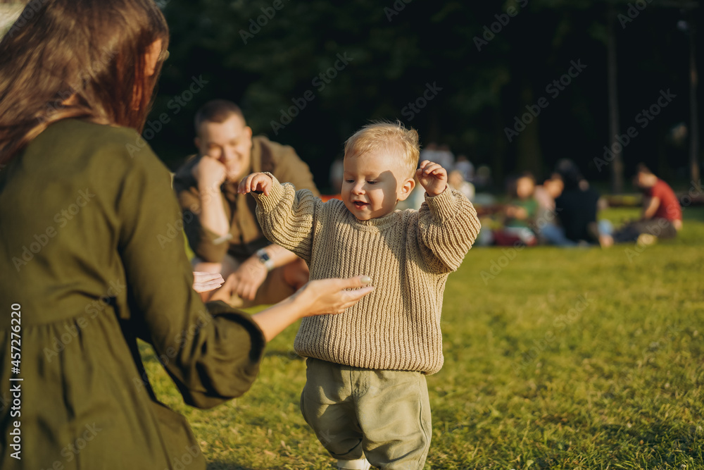 little caucasian boy learning to walk on lawn in park. His mother ...