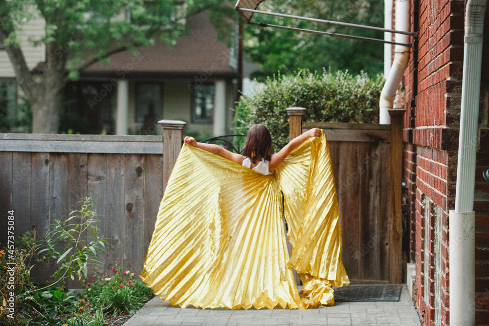 Rear-view of child in long golden cape with arms outstretched outside ...