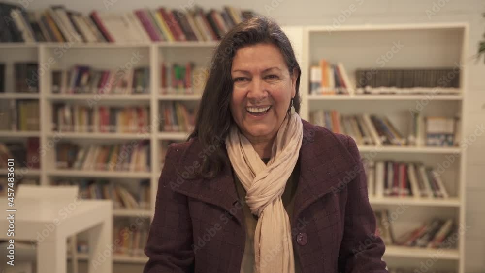 Beautiful Middle aged woman laughing in library out loud looking at camera stand indoors ...