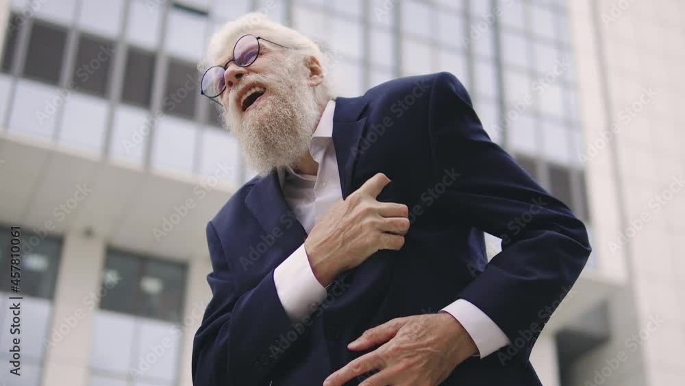 Elderly gray-haired man with long beard writhing in pain and screaming holding and massaging ...